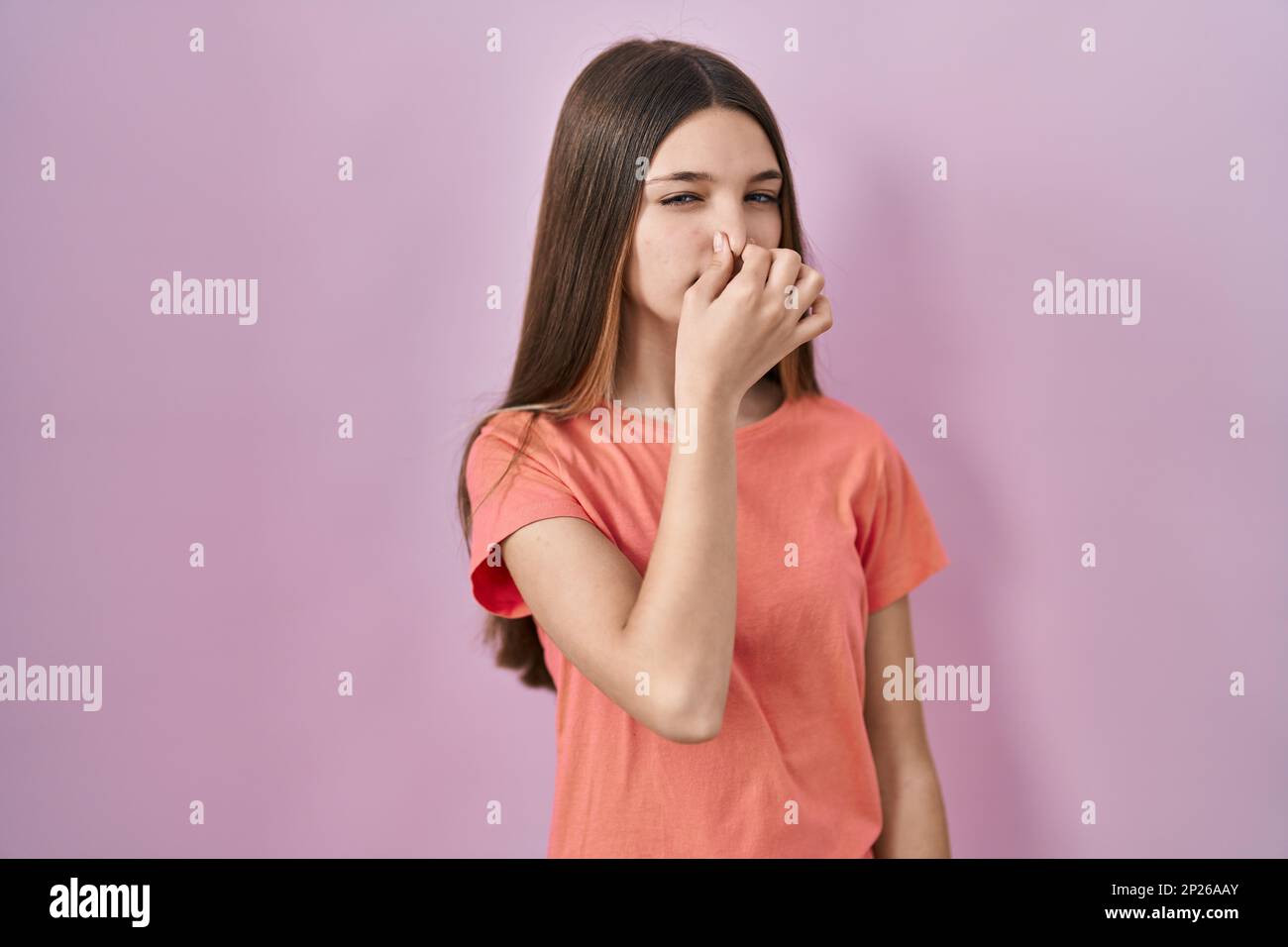 Teenager girl standing over pink background smelling something stinky ...