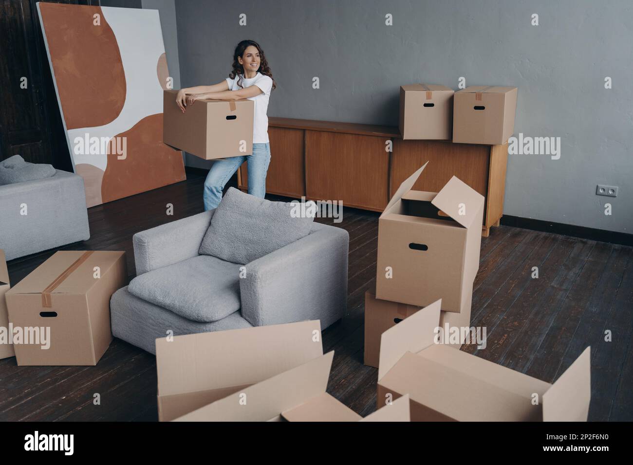 Happy female tenant holding box with personal things, standing in room ...