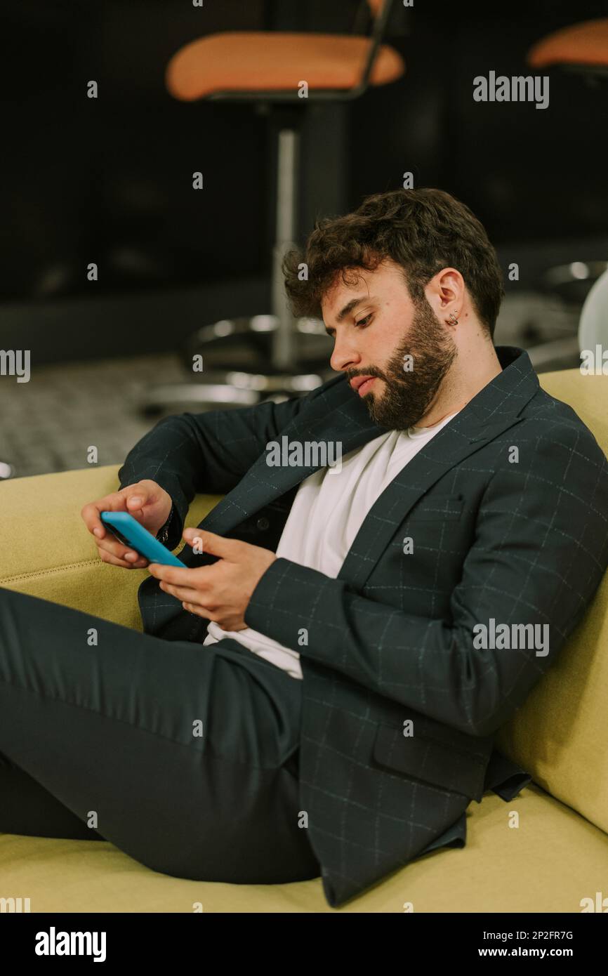 A tired good looking business person scrolling on his phone while lying on a yellow sofa at ...