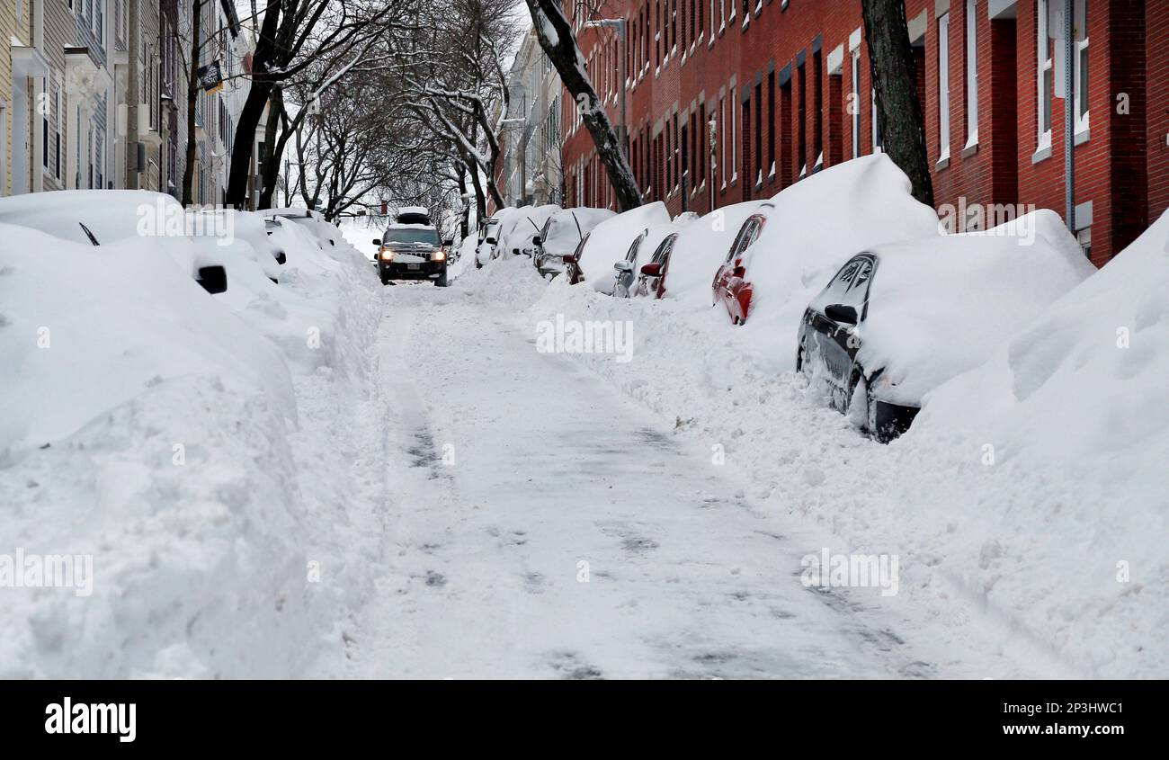 A car makes its way down a street filled with snowed-in vehicles in ...
