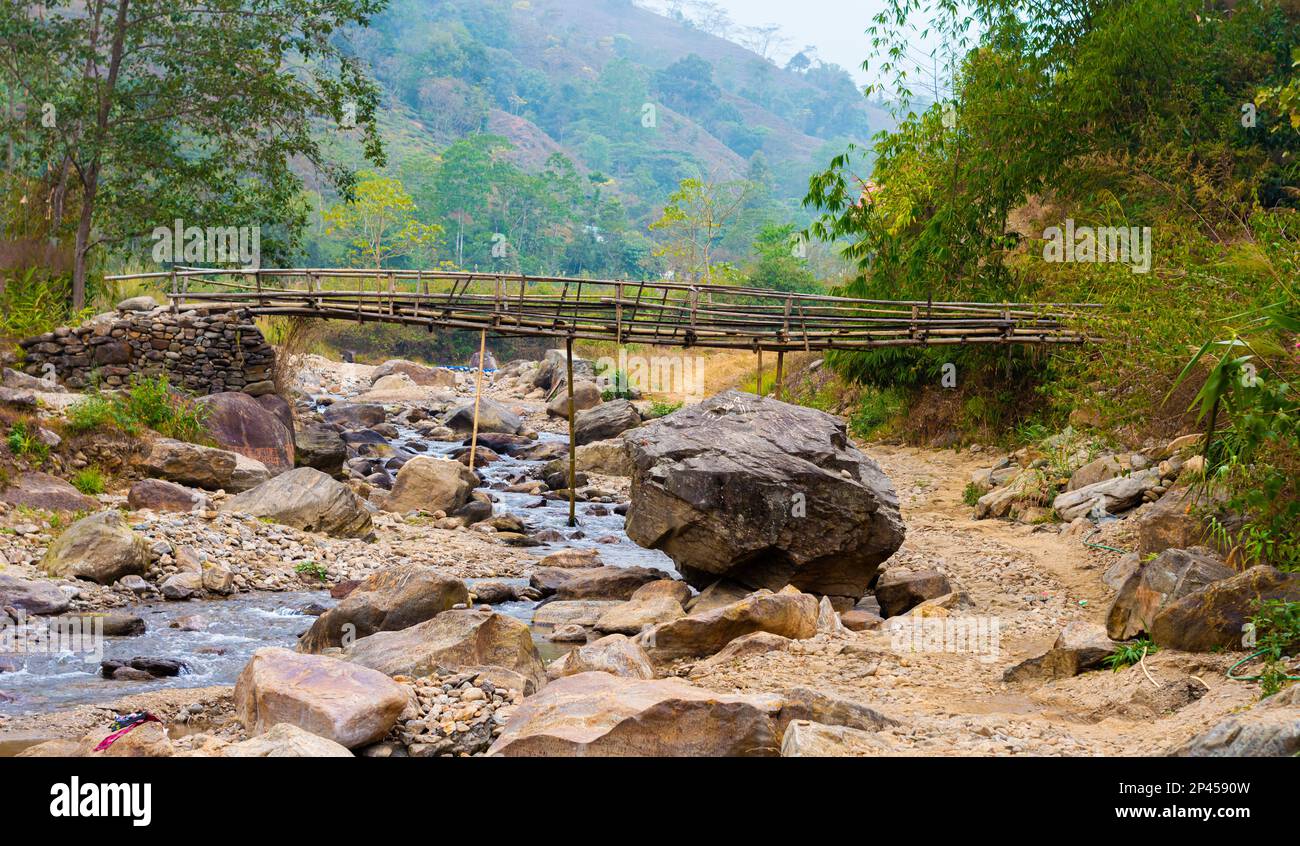A bamboo bridge over over a narrow rocky mountain river against a hill ...
