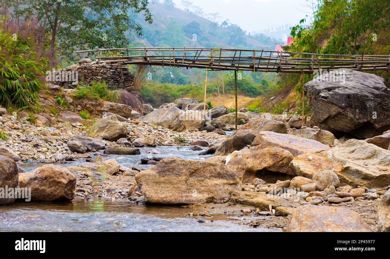 A bamboo bridge over over a narrow rocky mountain river against a hill ...