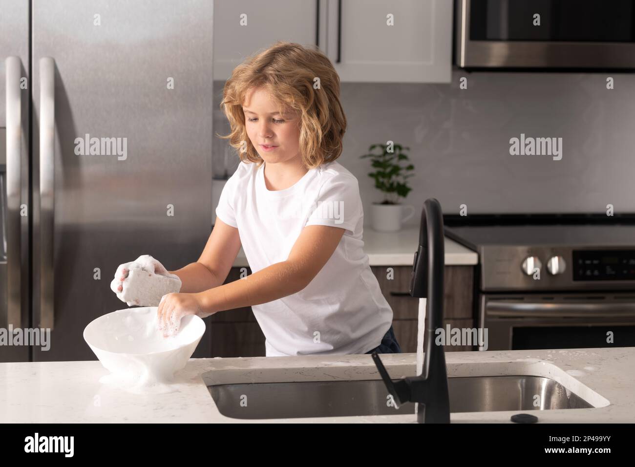 Kid boy washing dishes in the kitchen interior. Dishwashing liquid with ...
