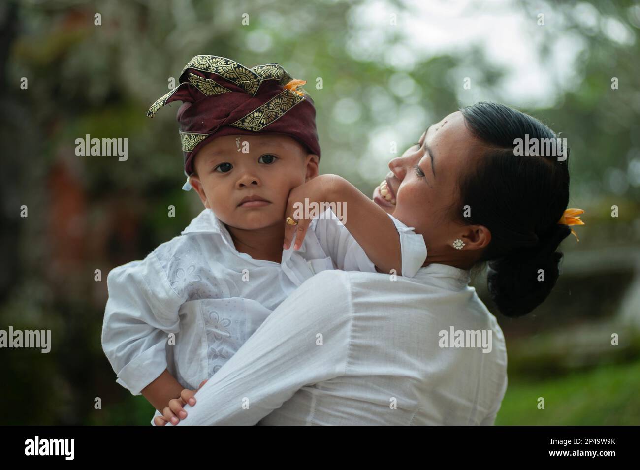 Boy wearing udeng (headress) in smiling mother's arms during Mepeed ...