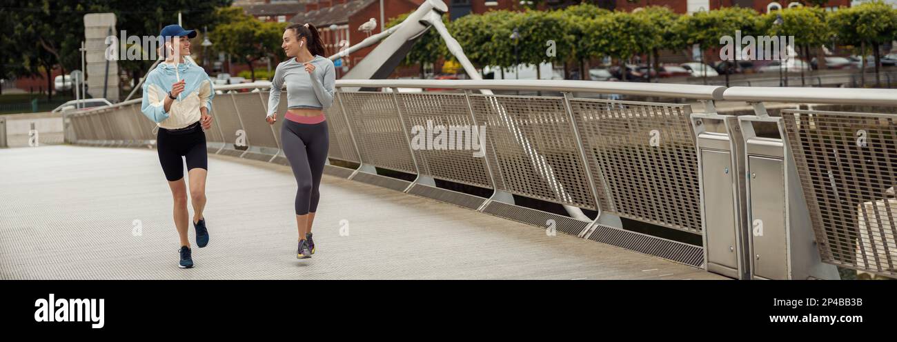 Two athletic women in sportswear is jogging on a bridge and listening ...