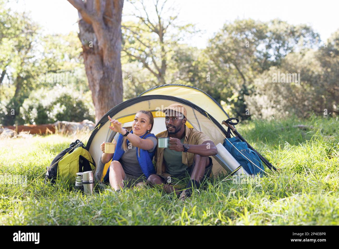 Happy, diverse couple in forest sitting by tent, drinking coffee and ...
