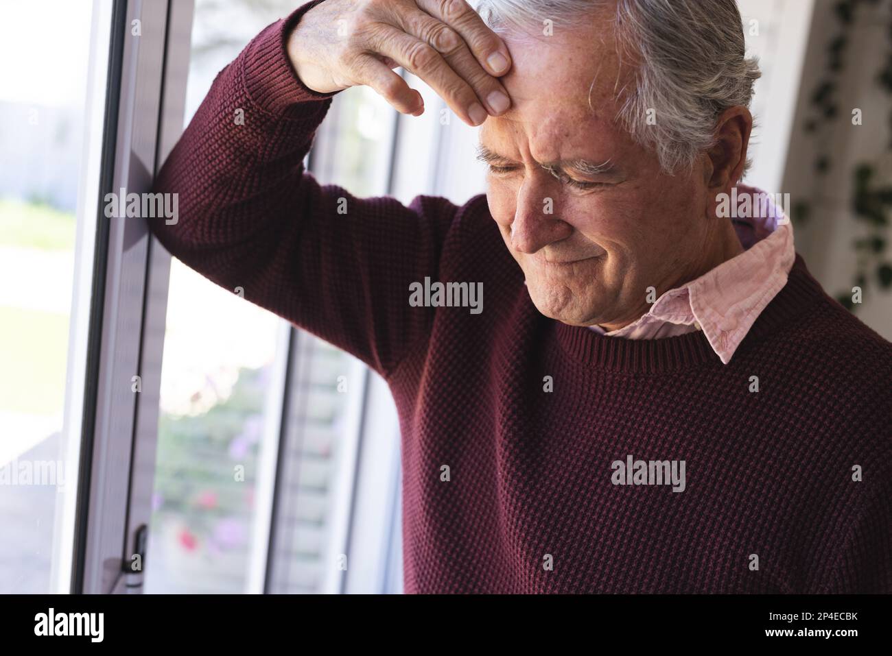 Distressed senior caucasian man at home leaning on window holding head ...