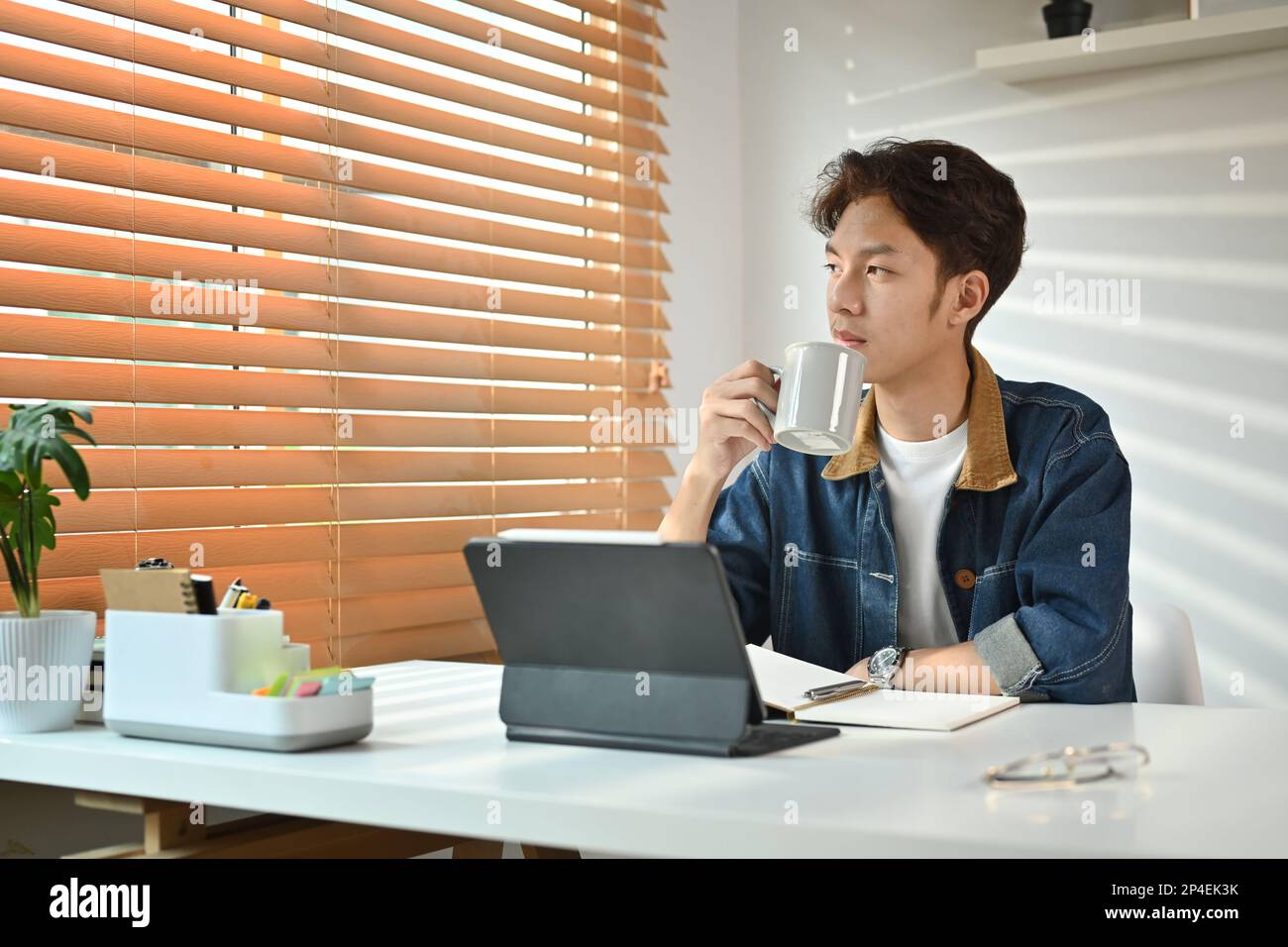 Thoughtful young asian man drinking coffee and looking through window ...