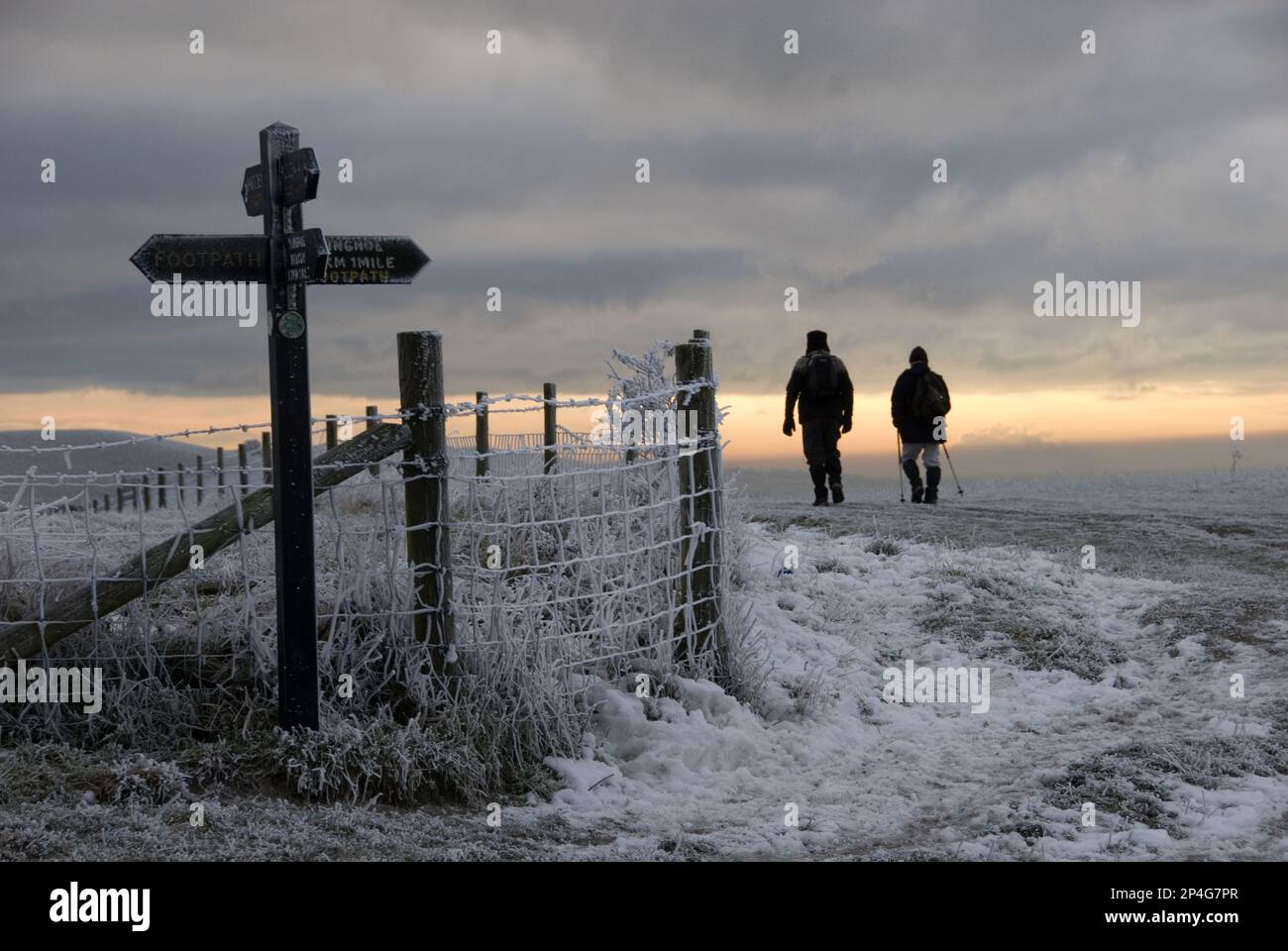 Walkers on frost and snow covered footpath, beside signpost and fence ...