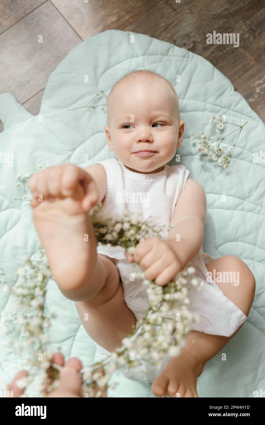 Bald baby girl in a white linen dress with gypsophila on a leaf rug ...