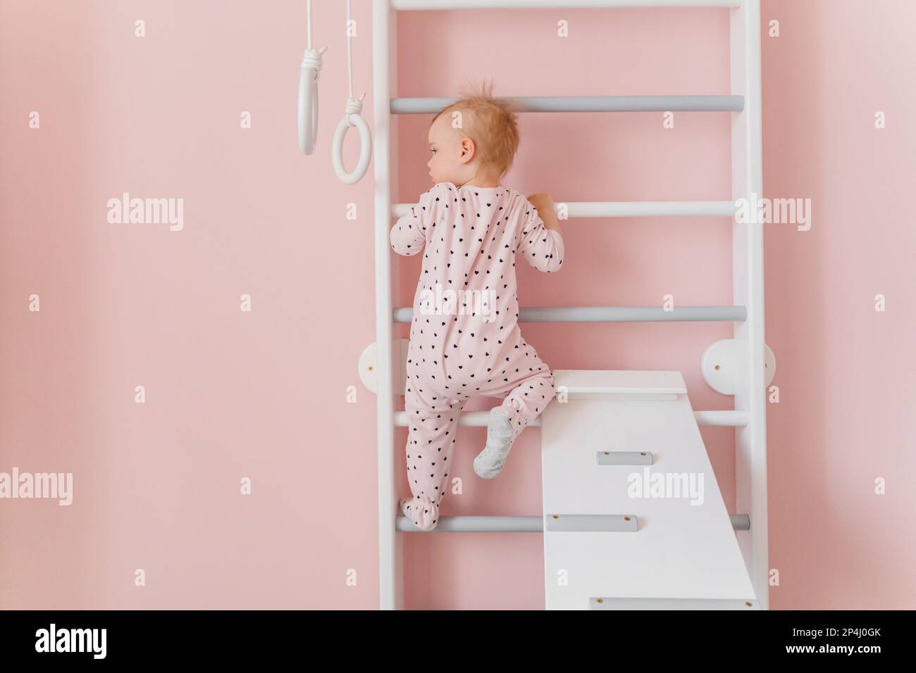 A cute toddler girl in soft pink pajamas does gymnastic on wall bars ...