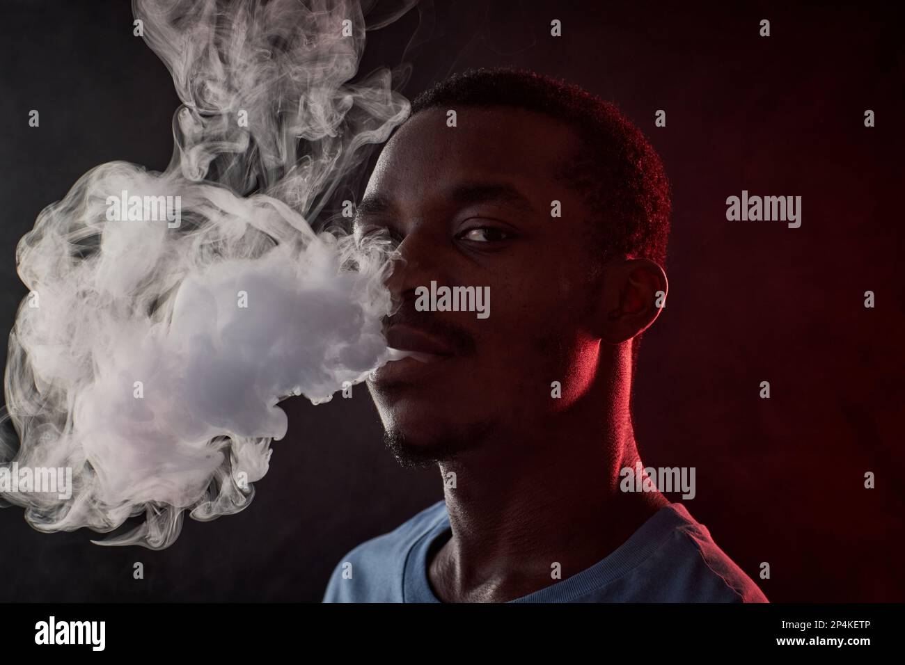 African American guy looking at camera while smoking in darkness and ...