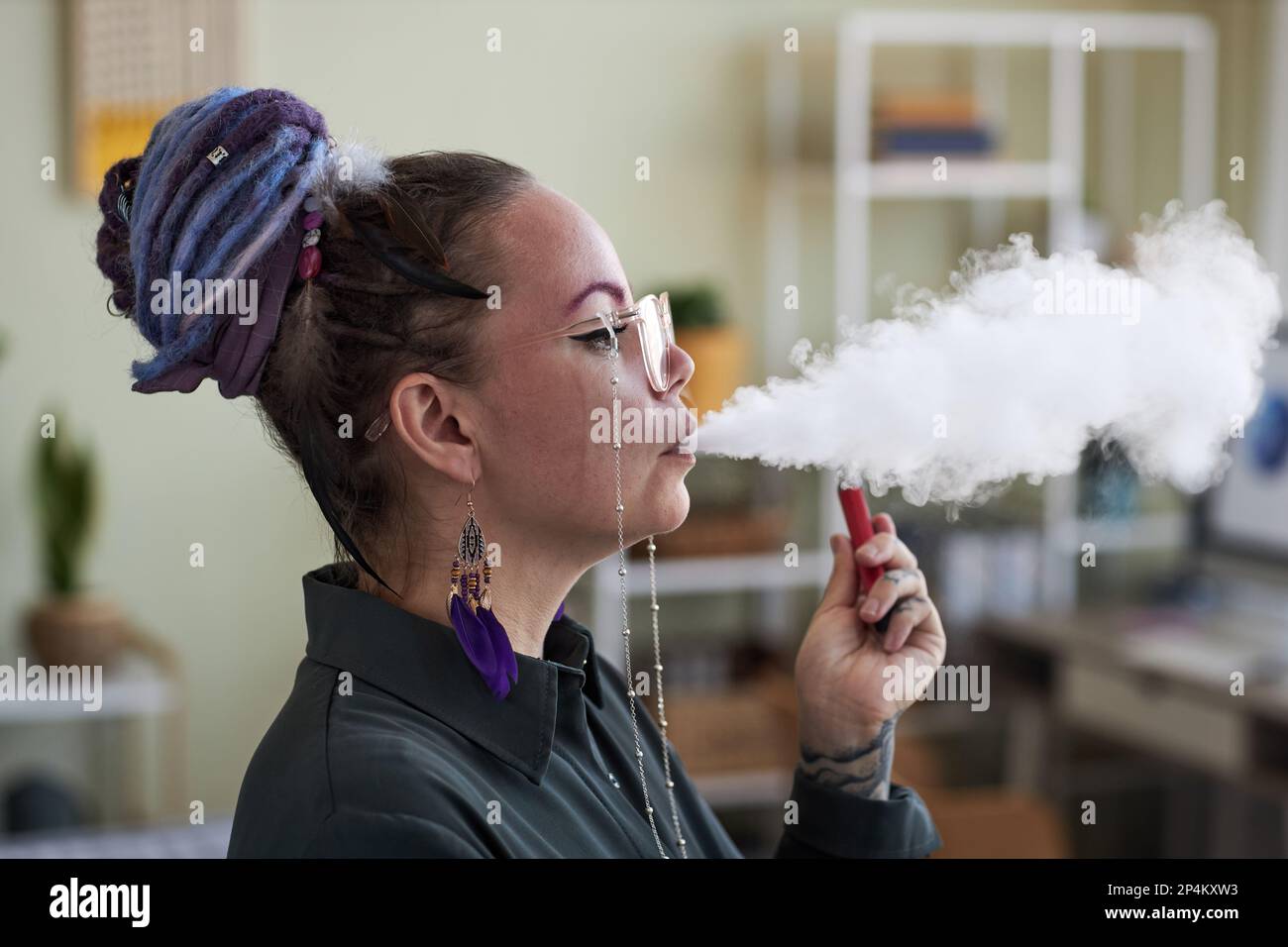 Side view of young woman in eyeglasses blowing white cloud of thick smoke out of her mouth while ...