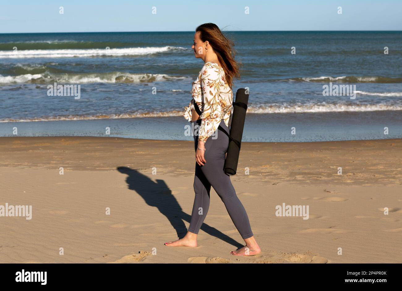 young woman walking barefoot on the beach with mat slung over her ...