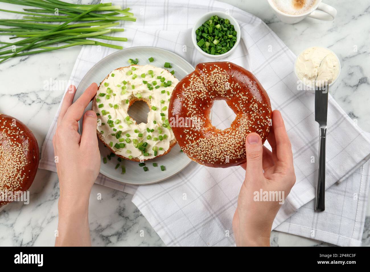 Woman making bagel with cream cheese and green onion at white marble ...