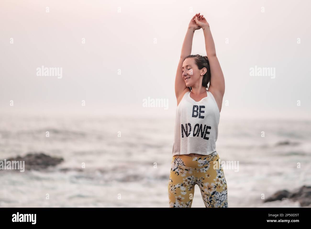 A female adult practising yoga poses on a rocky beach, balancing on a ...
