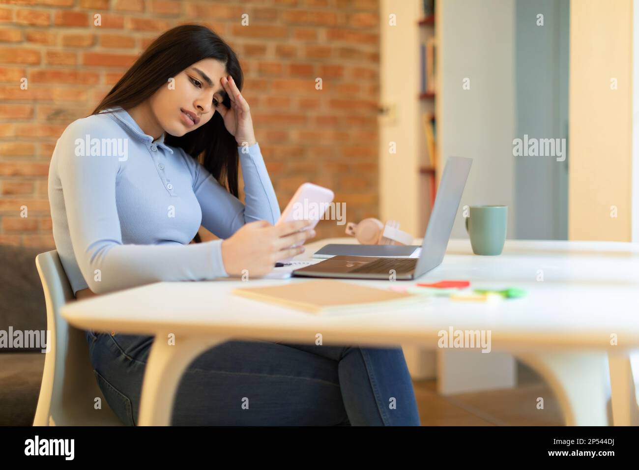 Unhappy bored teen lady sitting at desk with laptop, using smartphone ...