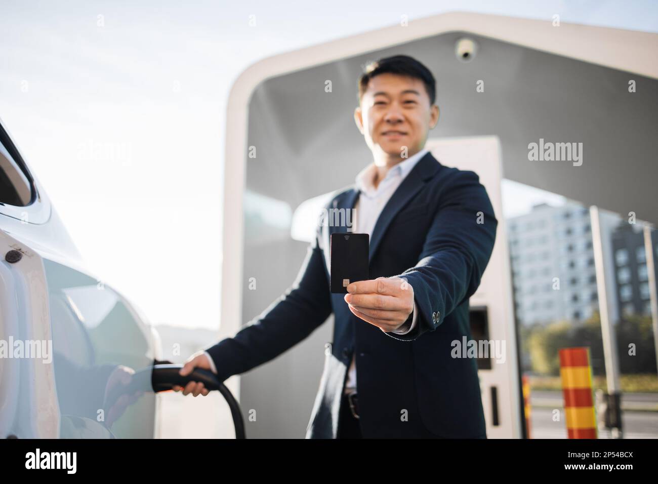 Positive asian man in stylish black suit charging white electric car ...