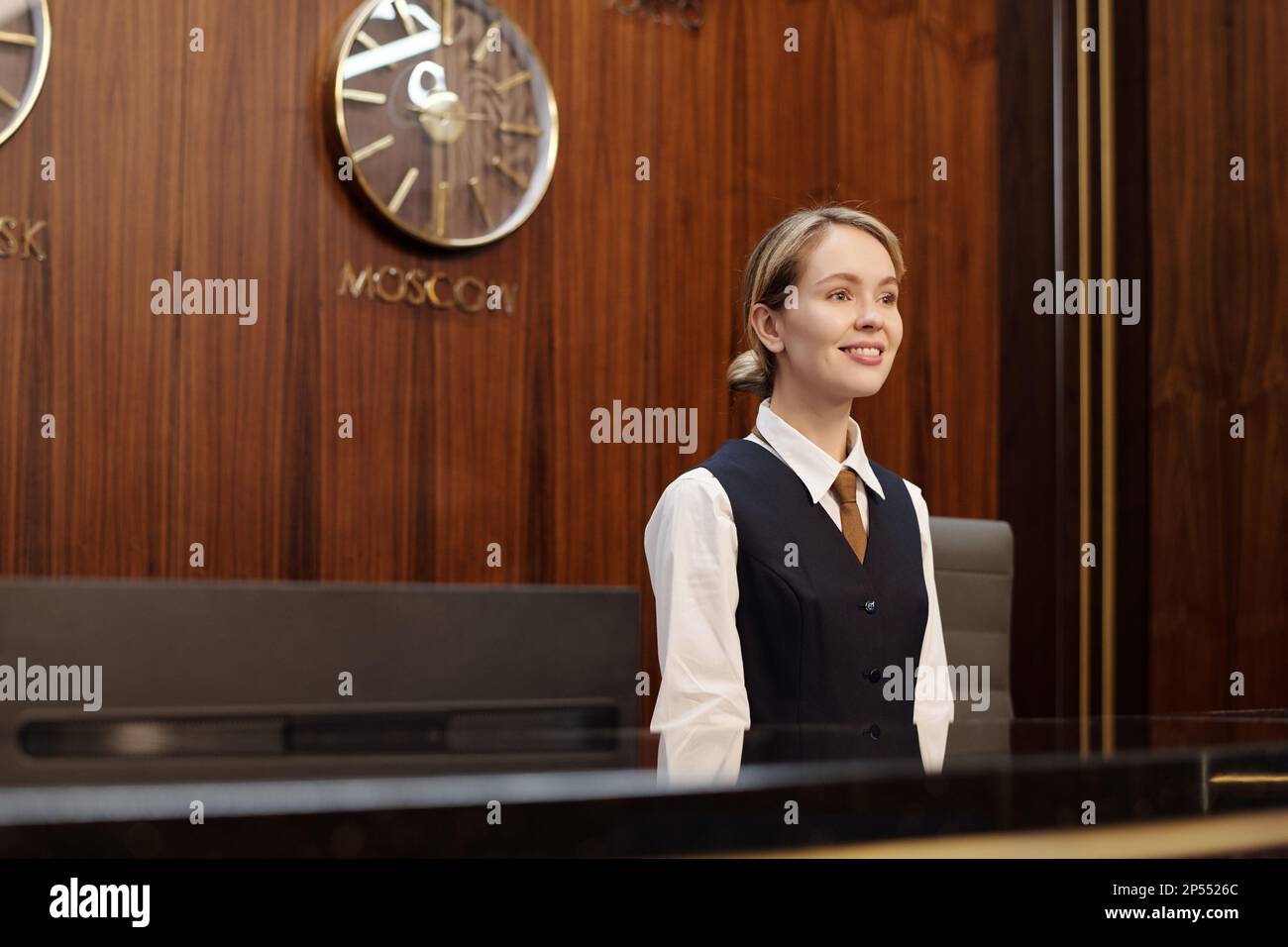 Young successful female receptionist in uniform standing by check-in ...