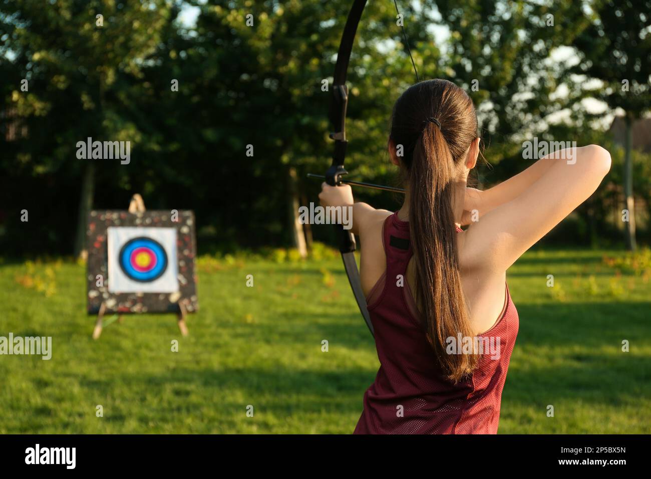 Woman with bow and arrow aiming at archery target in park, back view ...