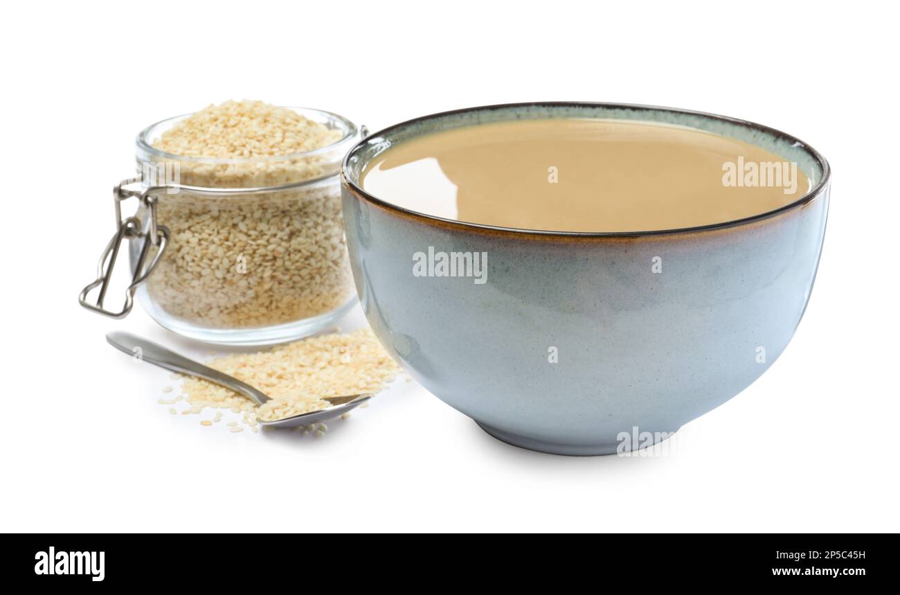 Bowl of tasty sesame paste, jar, spoon and seeds on white background ...