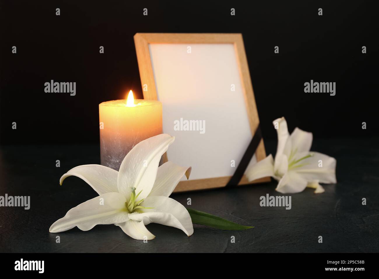 Blank funeral frame, burning candle and lily flowers on dark table ...
