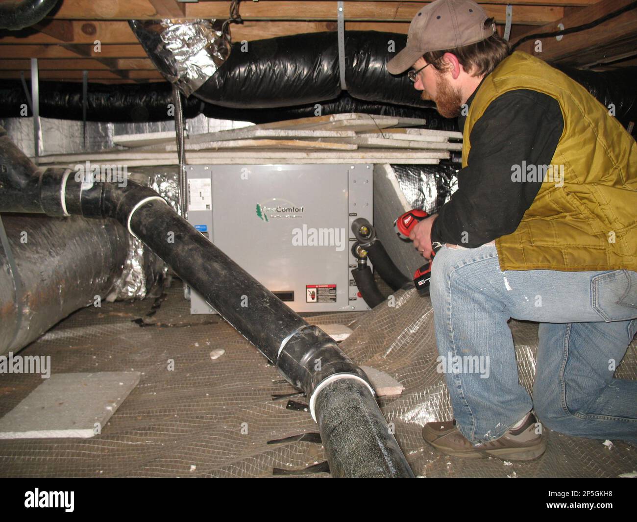 John Kajdan kneels in his crawl space in front of the heat pump, a ...