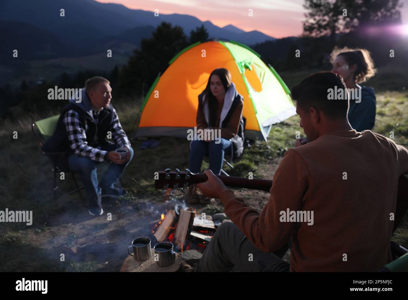 Group of friends gathering around bonfire at camping site in evening ...