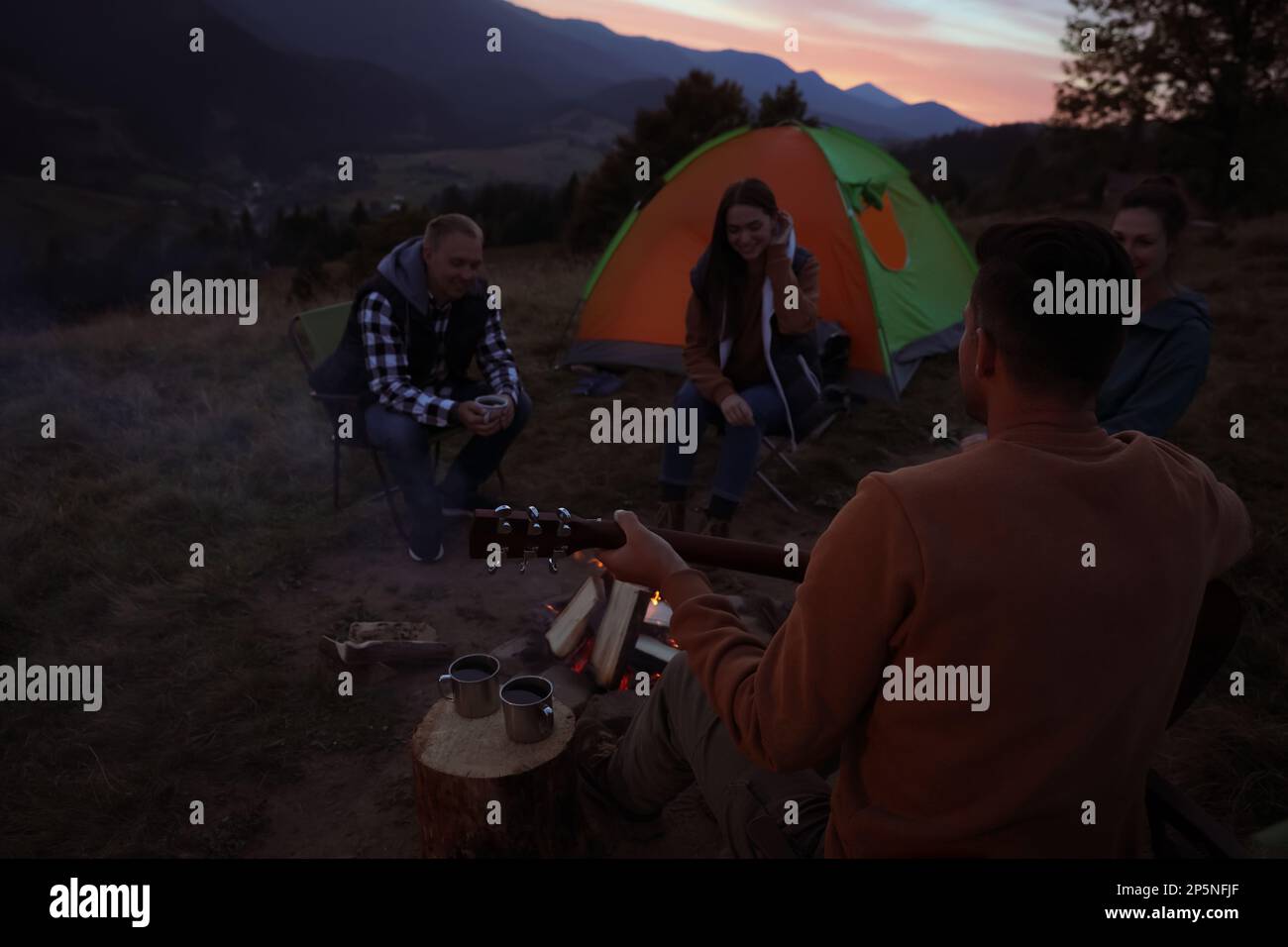Group of friends gathering around bonfire at camping site in evening ...