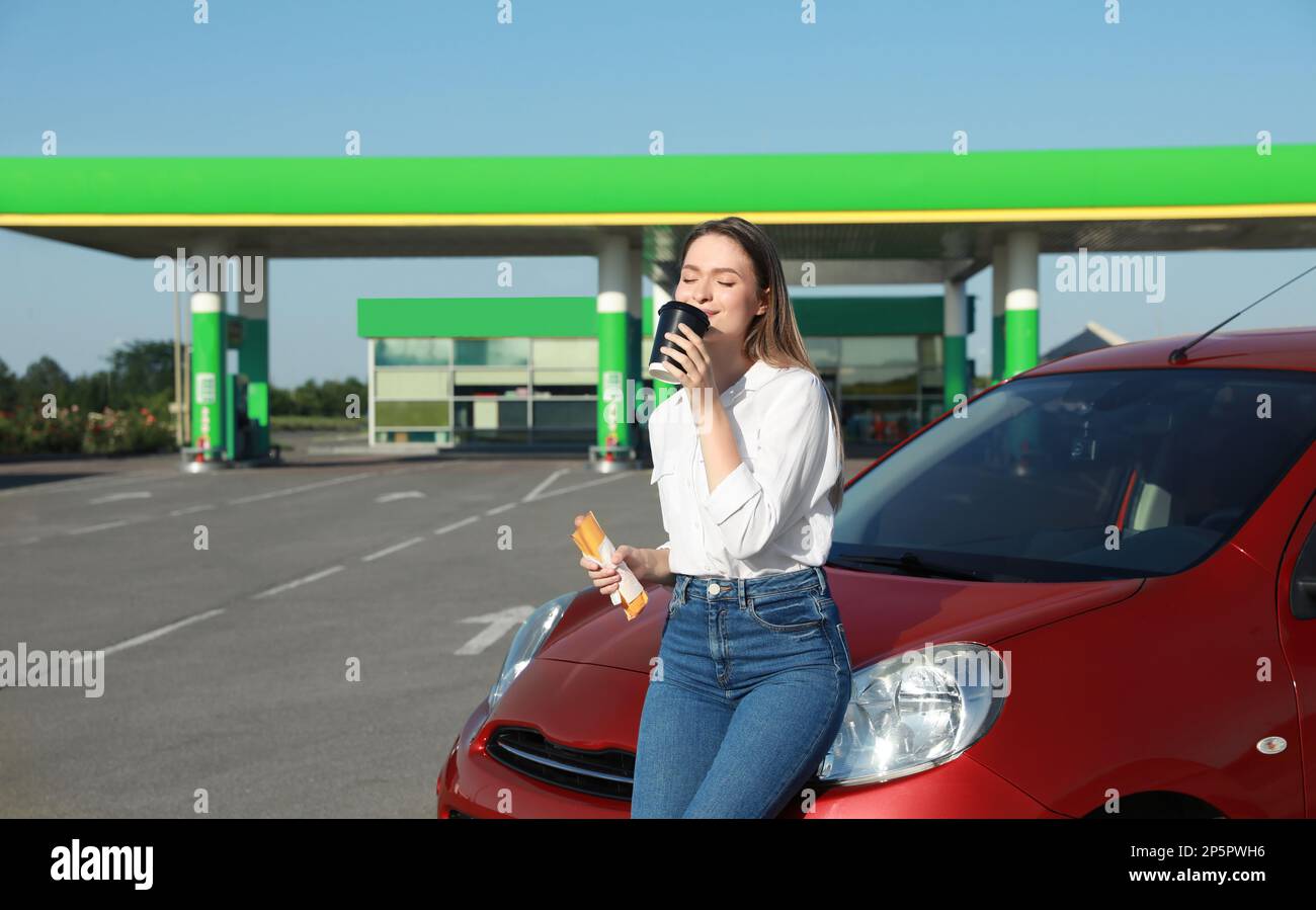 Beautiful young woman with hot dog drinking coffee near car at gas ...