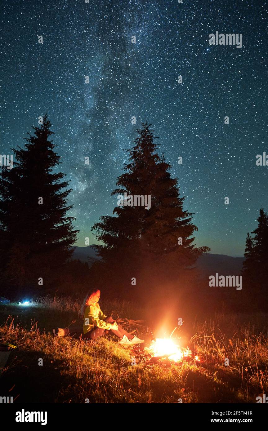 Night camping in mountains under starry sky with Milky way. Woman sitting on grass near campfire ...