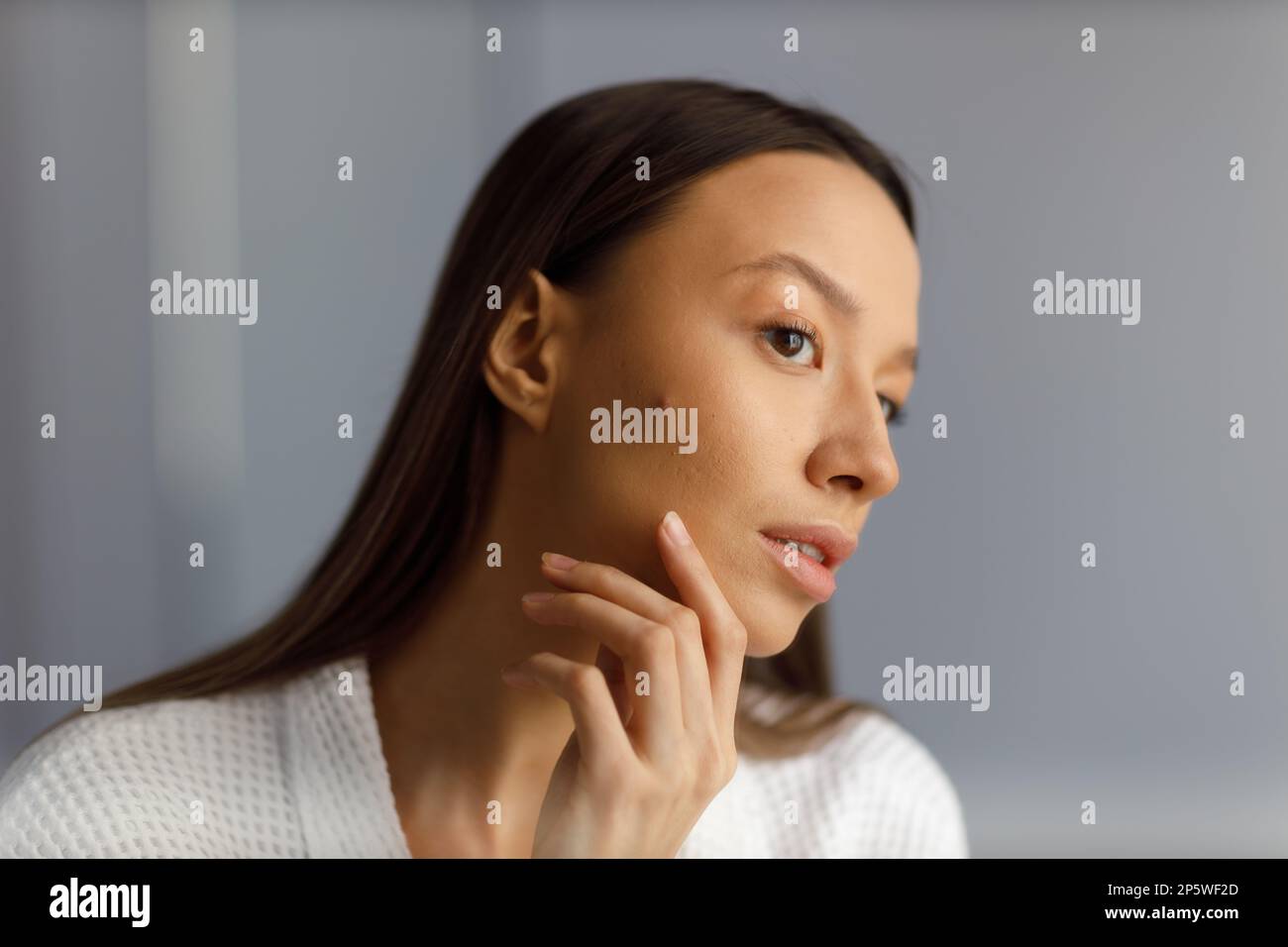 Attractive upset young woman in white bathrobe examines pimples on her ...
