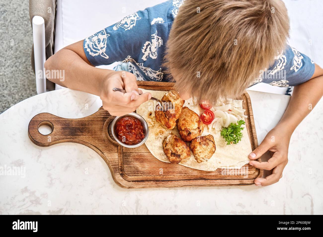 Blond boy eats grilled meat dish sitting at table in restaurant close ...
