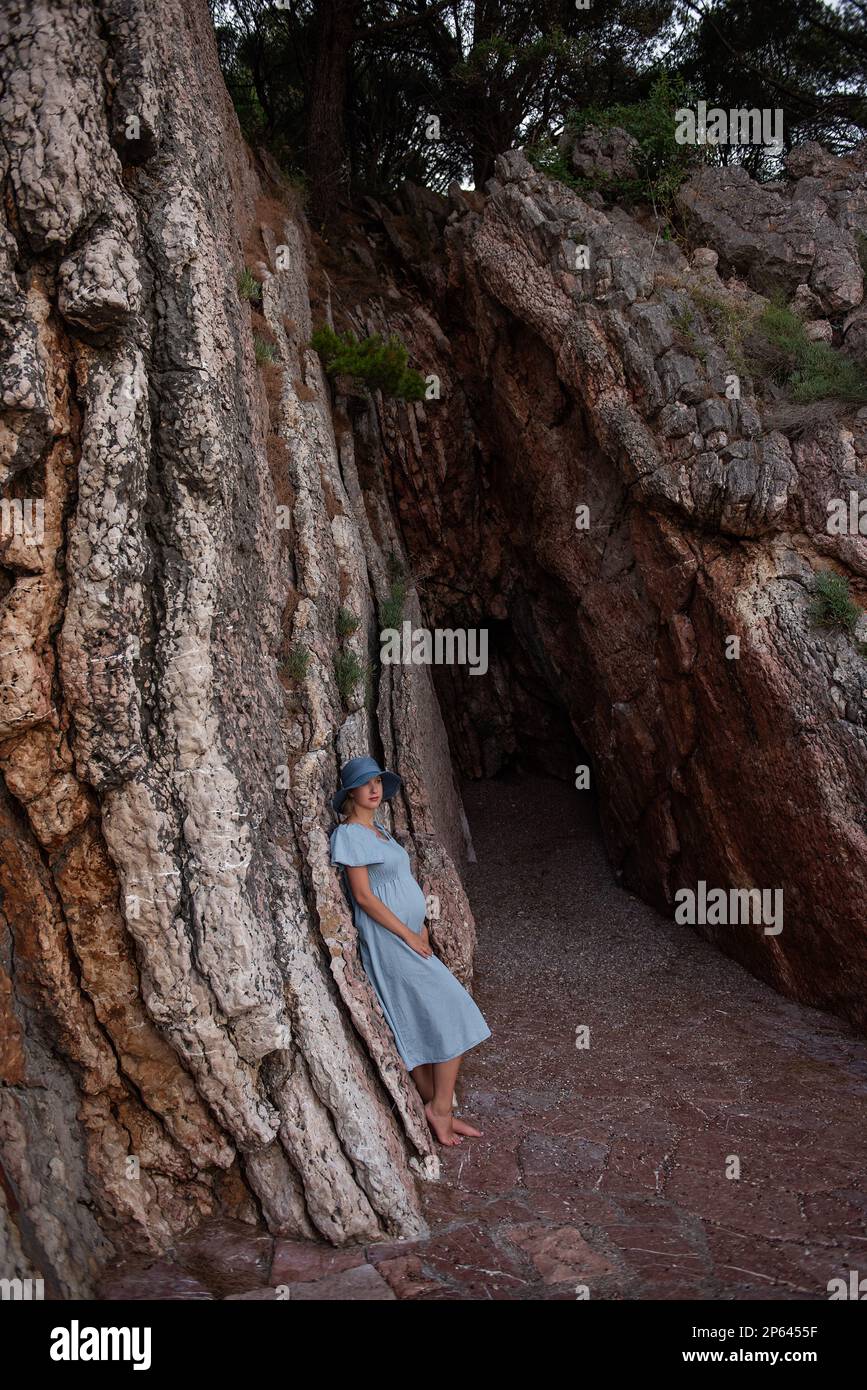Pregnant millennial woman in blue dress stands among the rocks on the ...