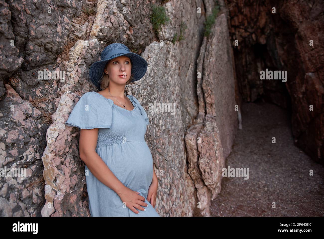 Pregnant millennial woman in blue dress stands among the rocks on the ...