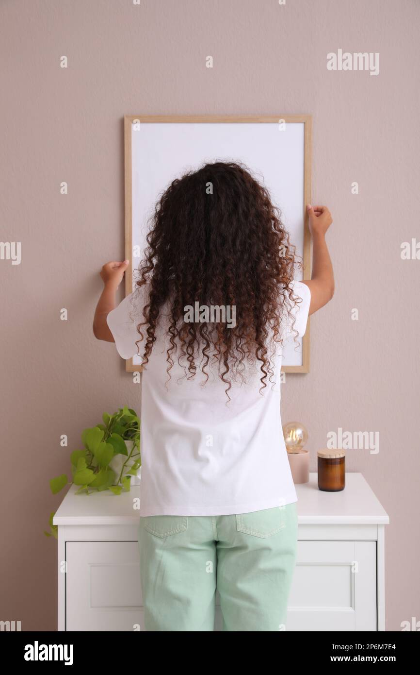 Woman hanging frame on pale rose wall over chest of drawers in room ...