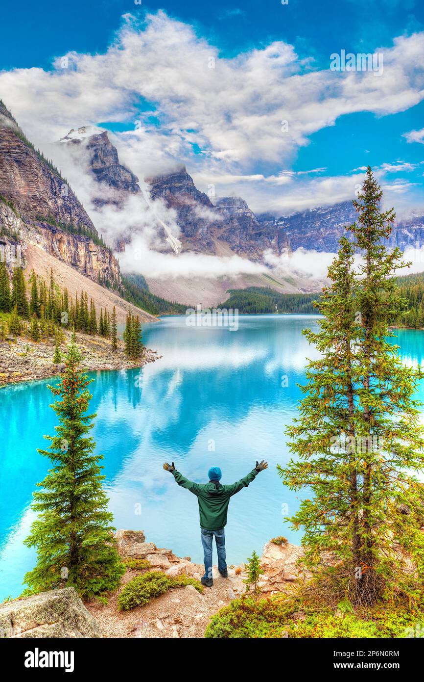 Hiker with arms raised at Moraine Lake as low clouds descend over the ...