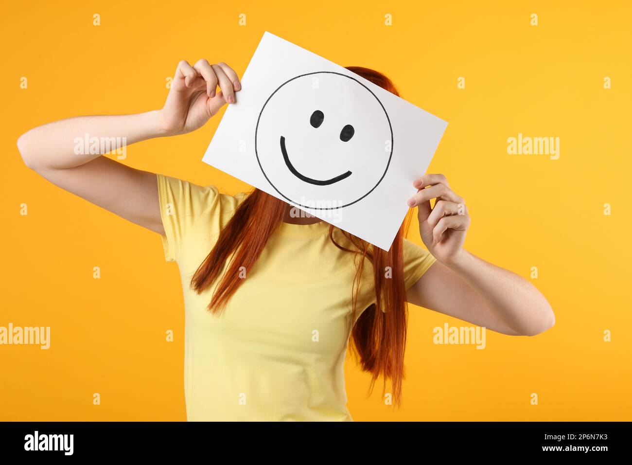 Woman hiding behind sheet of paper with happy face on yellow background ...