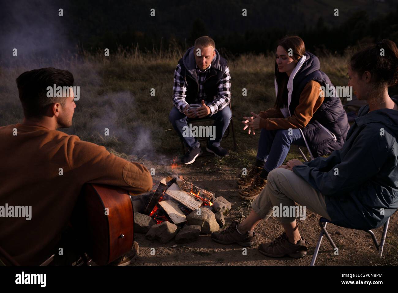 Group of friends gathering around bonfire at camping site in evening ...