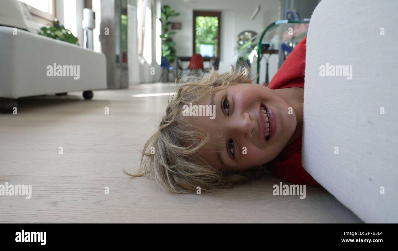 One happy small boy lying on floor hiding behind couch at modern home ...