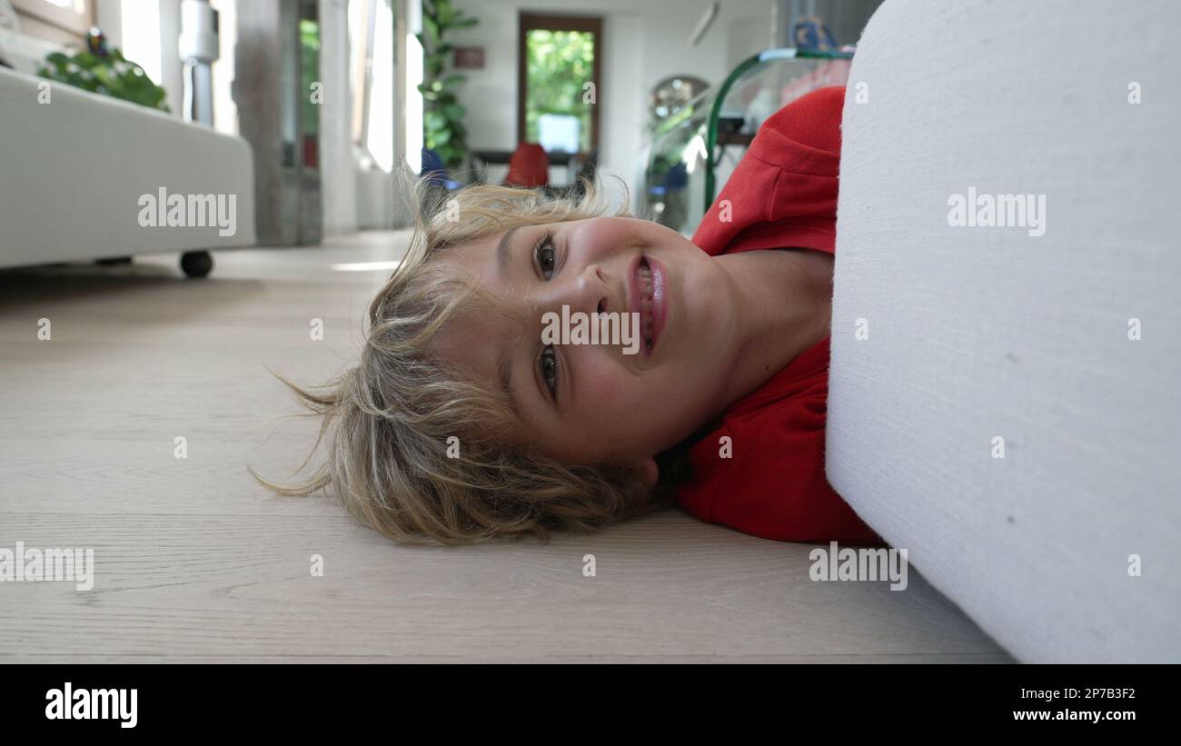 One happy small boy lying on floor hiding behind couch at modern home ...