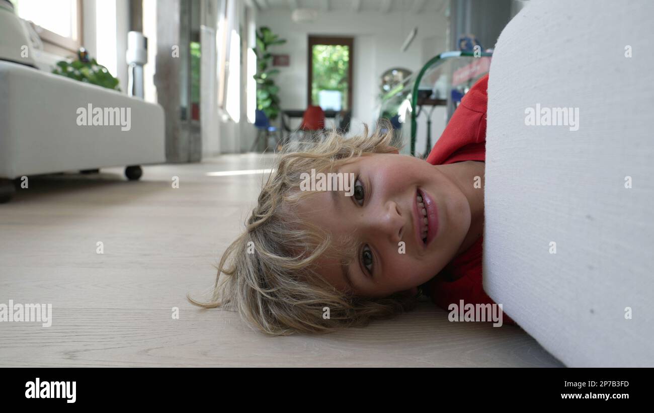 One happy small boy lying on floor hiding behind couch at modern home ...