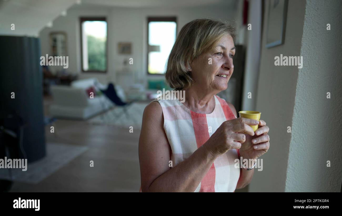 Senior woman drinking coffee in the morning standing by window at home ...