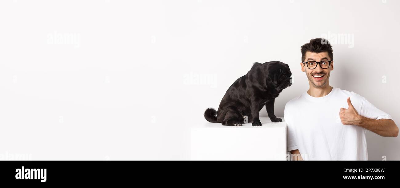 Image of happy and satisfied young man sitting near pug dog and showing ...