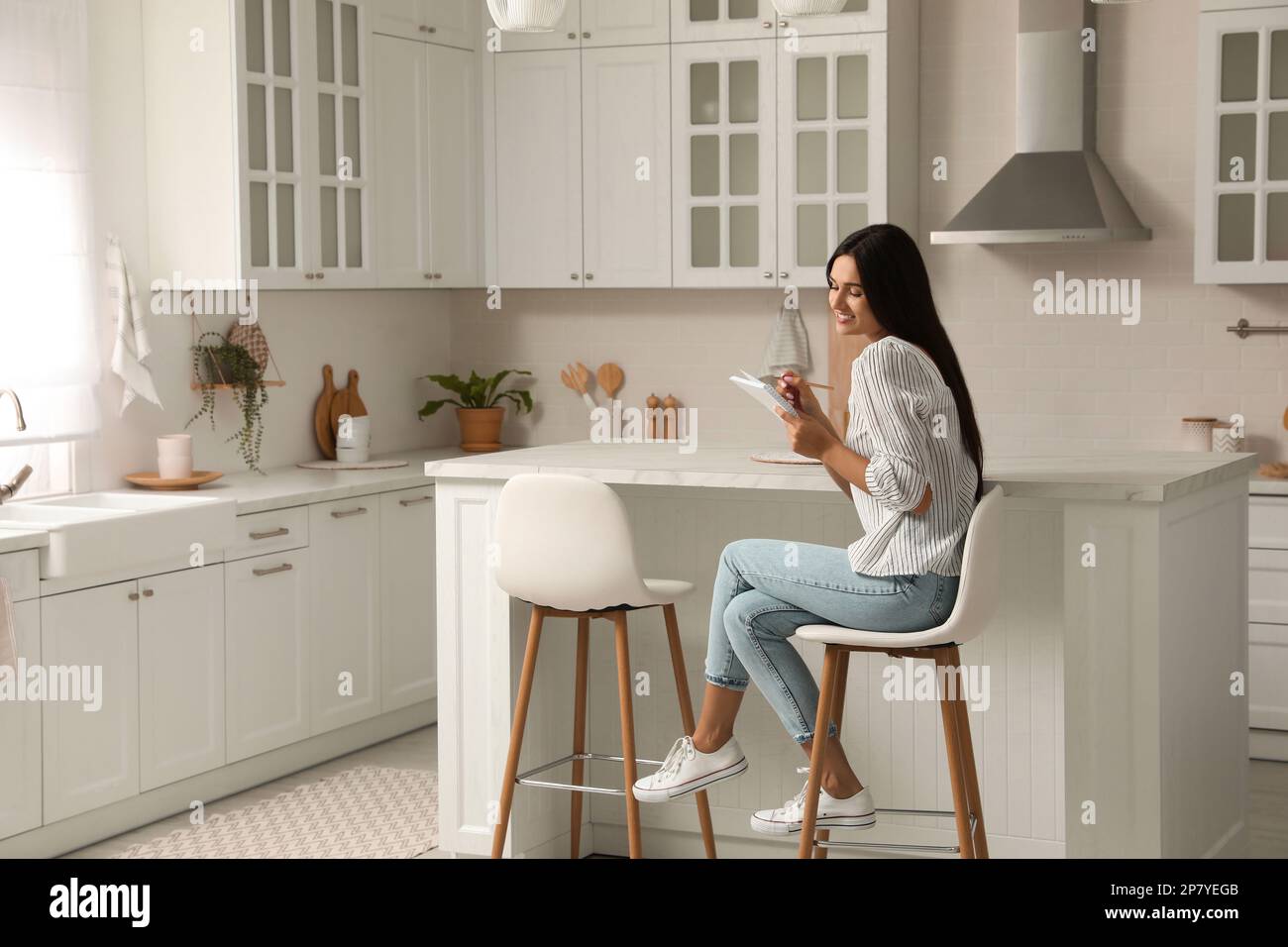 Beautiful young woman with notebook sitting on stool in kitchen Stock ...