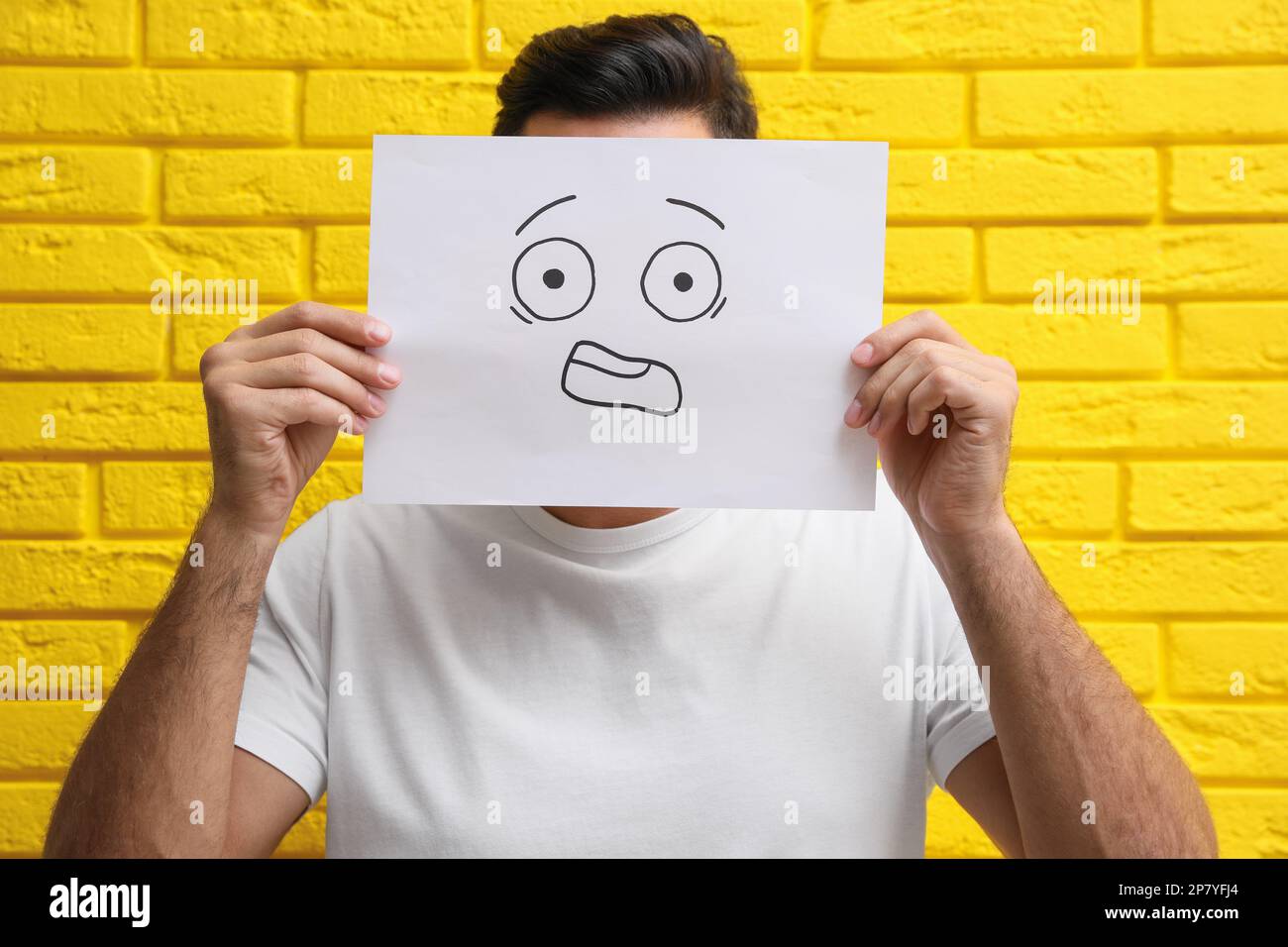 Man hiding emotions using card with drawn scared face near yellow brick ...