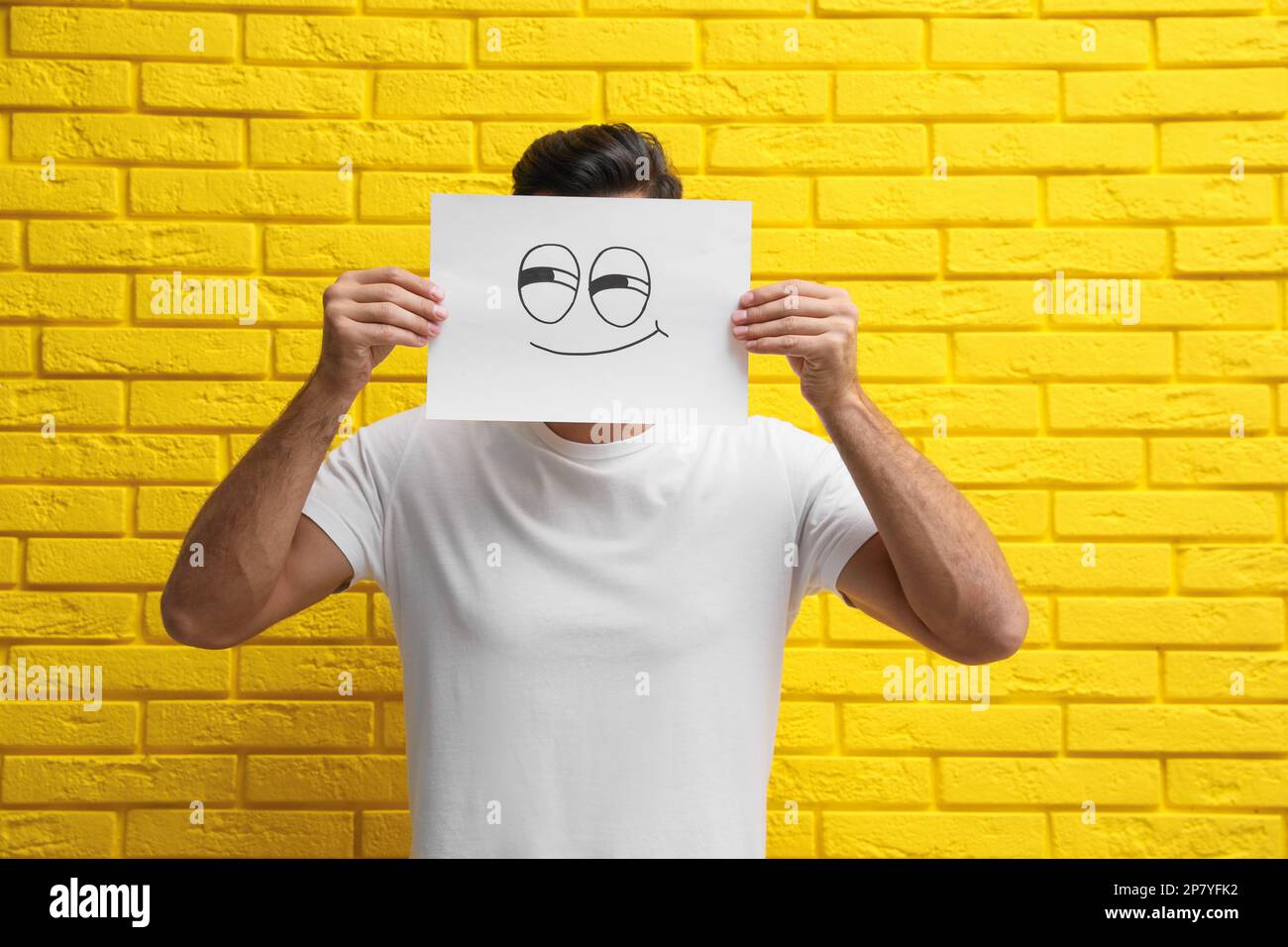 Man hiding emotions using card with drawn smiling face near yellow ...