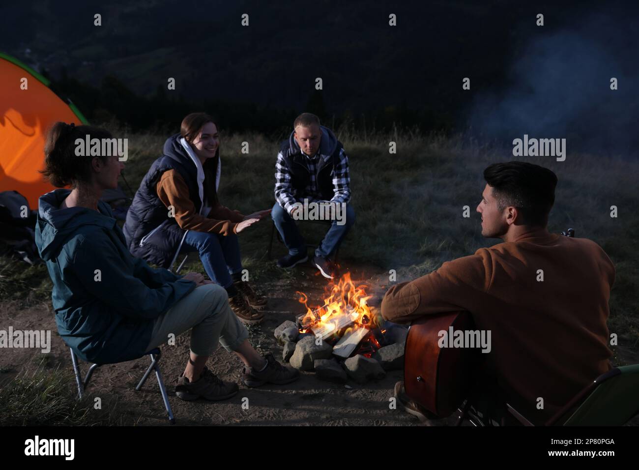 Group of friends gathering around bonfire at camping site in evening ...