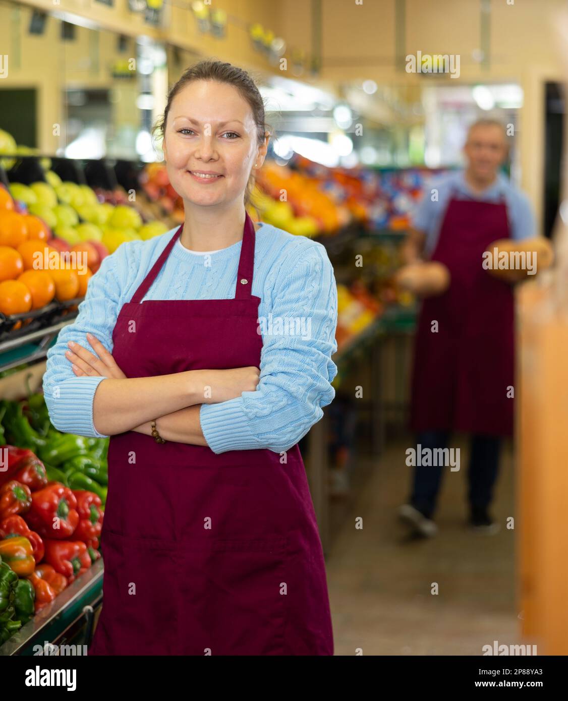 Cheerful middle-aged female supermarket worker in apron standing in ...