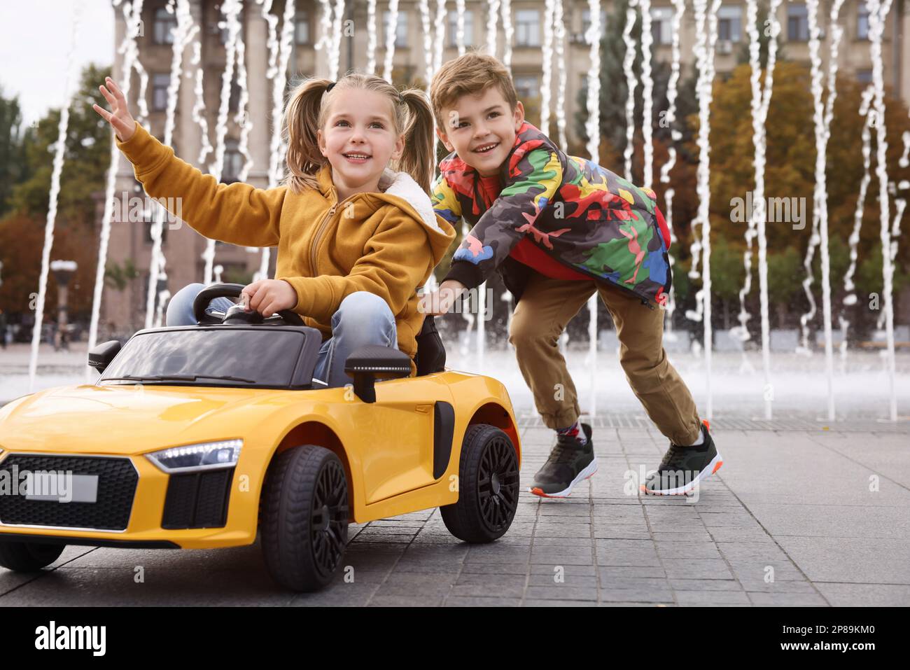 Cute boy pushing children's car with little girl near fountain on city ...